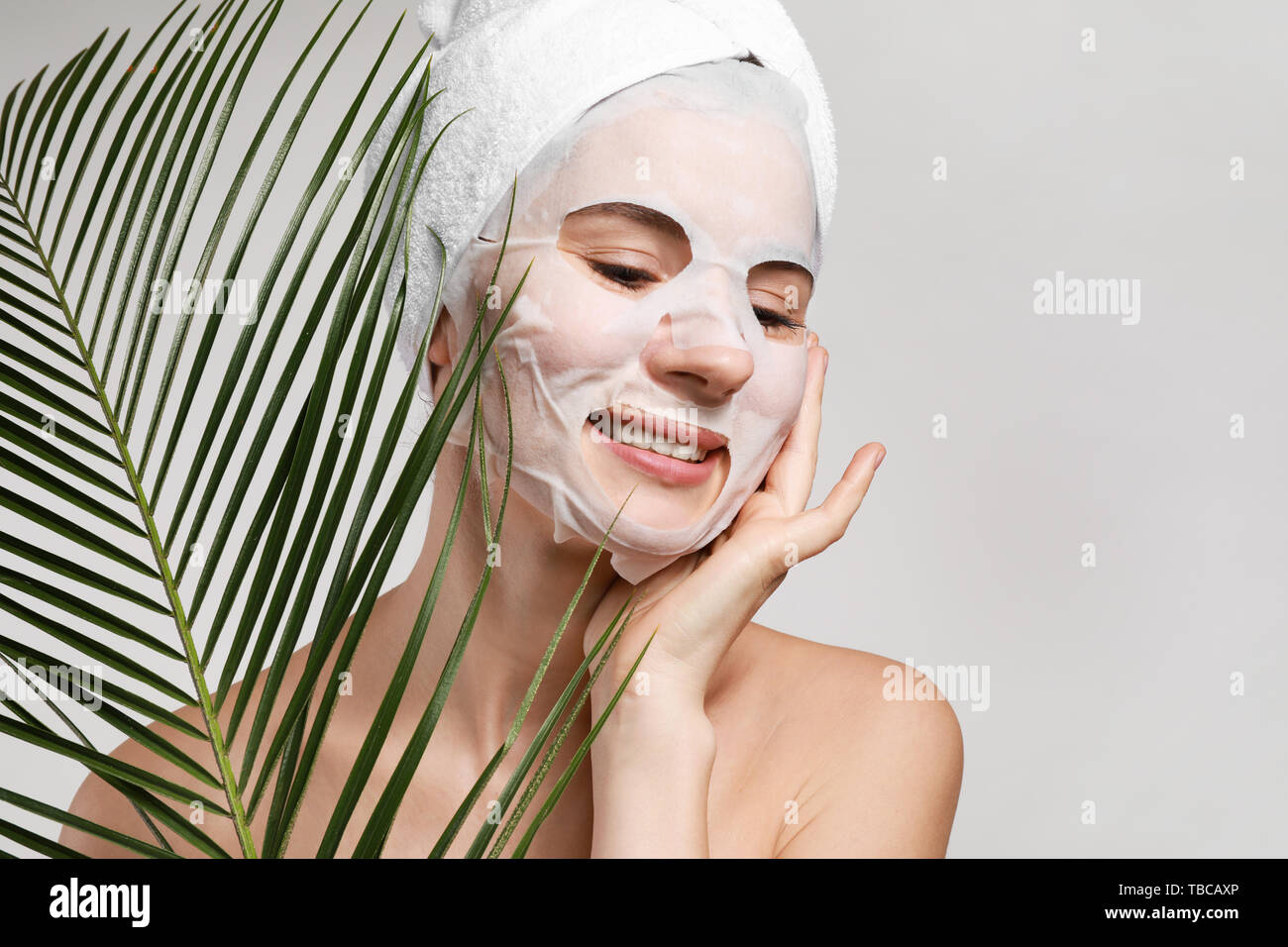 Woman with sheet facial mask and tropical leaf on light background ...