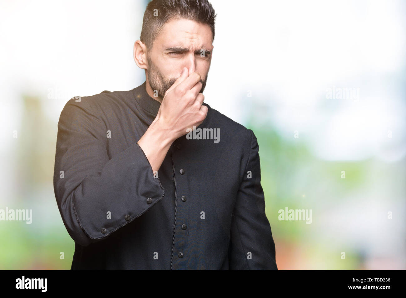 Young Christian priest over isolated background smelling something ...