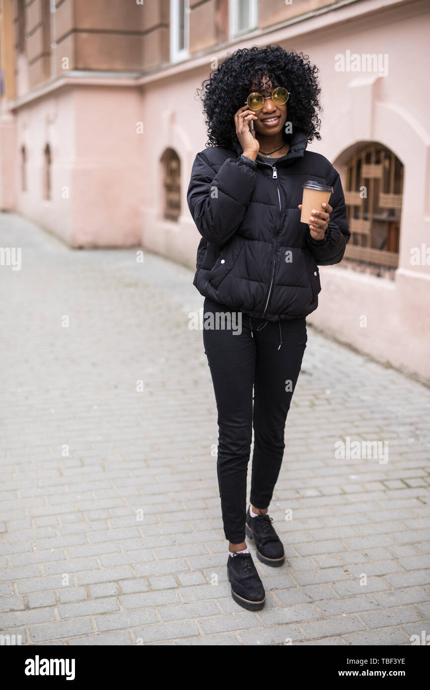 Cheerful Young African Woman Wearing Coat Walking Outdoors Holding