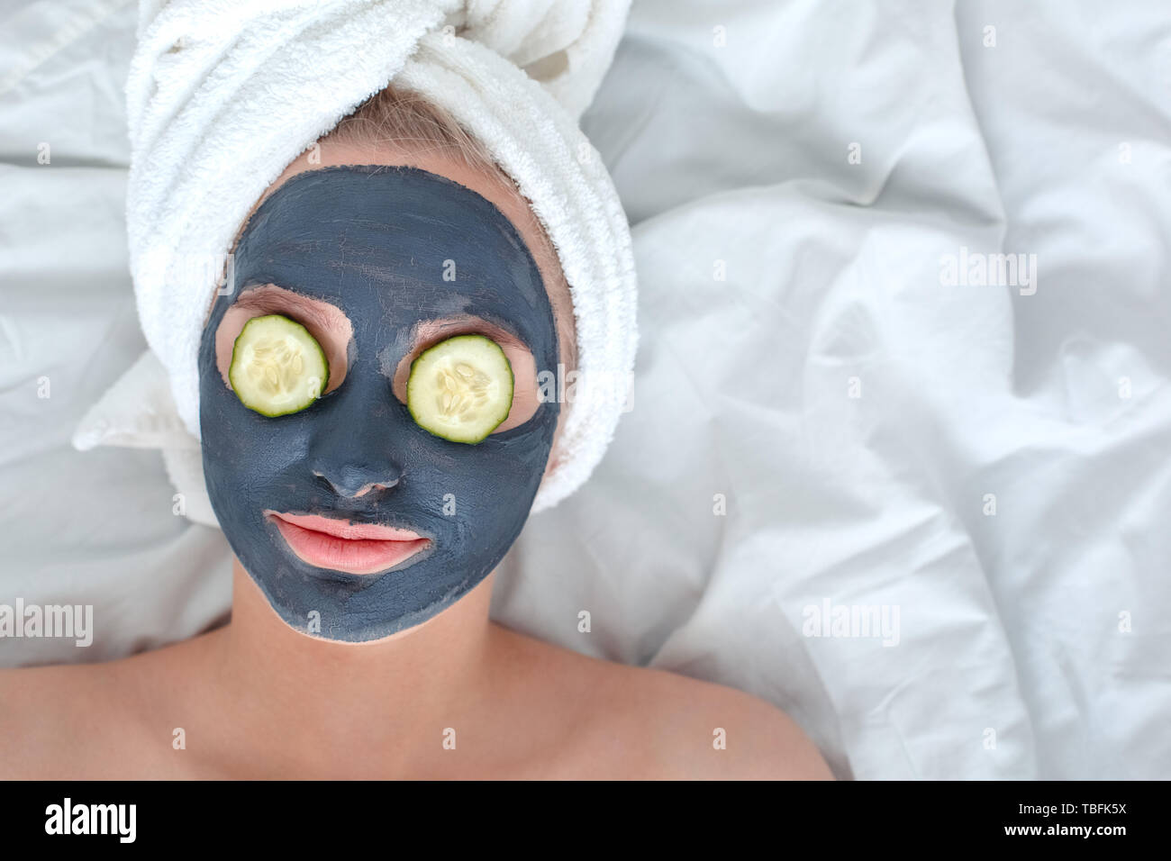 Young woman rest at home beauty care lying on bed with a black clay mask and cucumber slices on ...