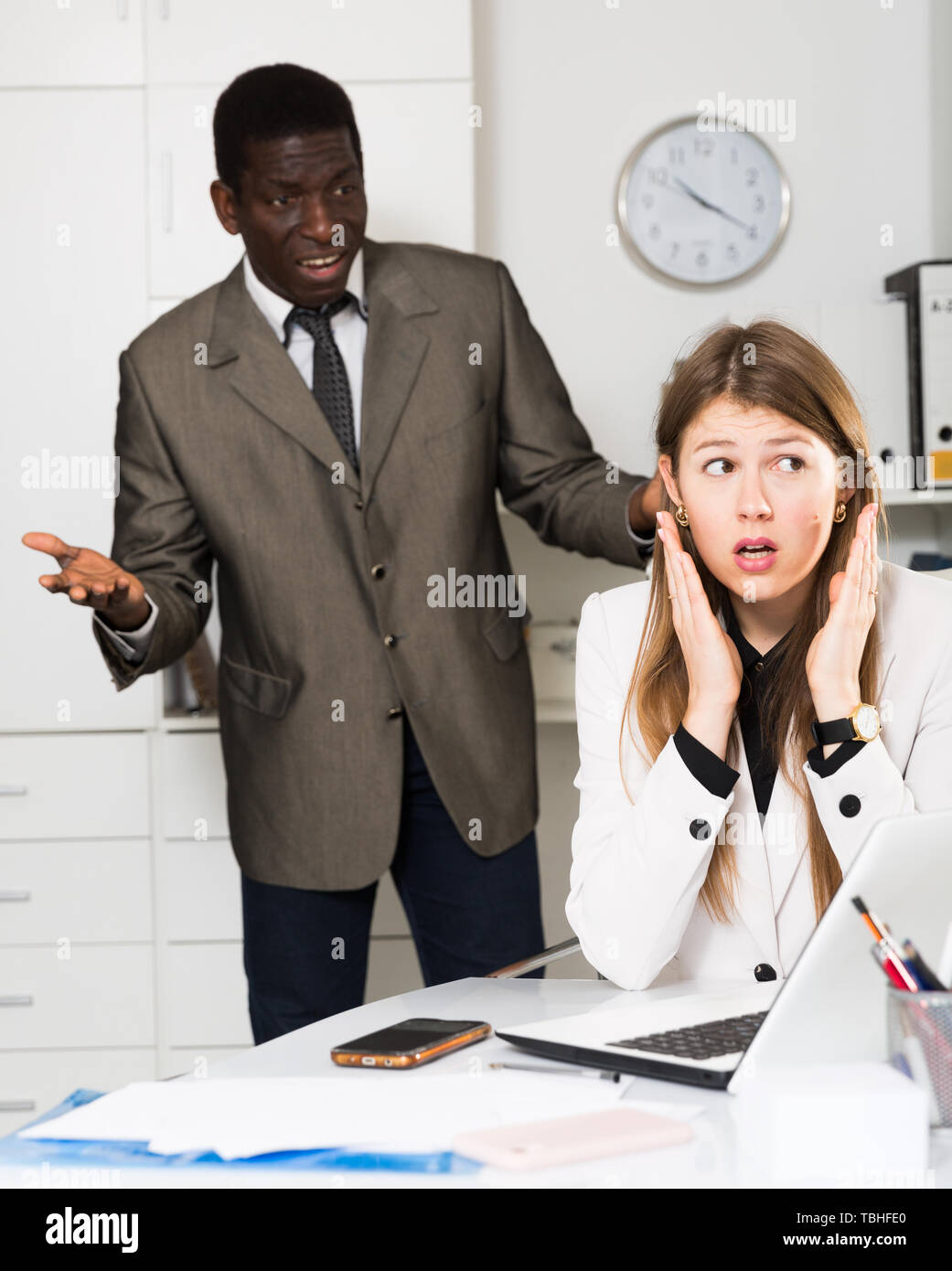 Frustrated woman sitting at office desk with disgruntled boss behind ...