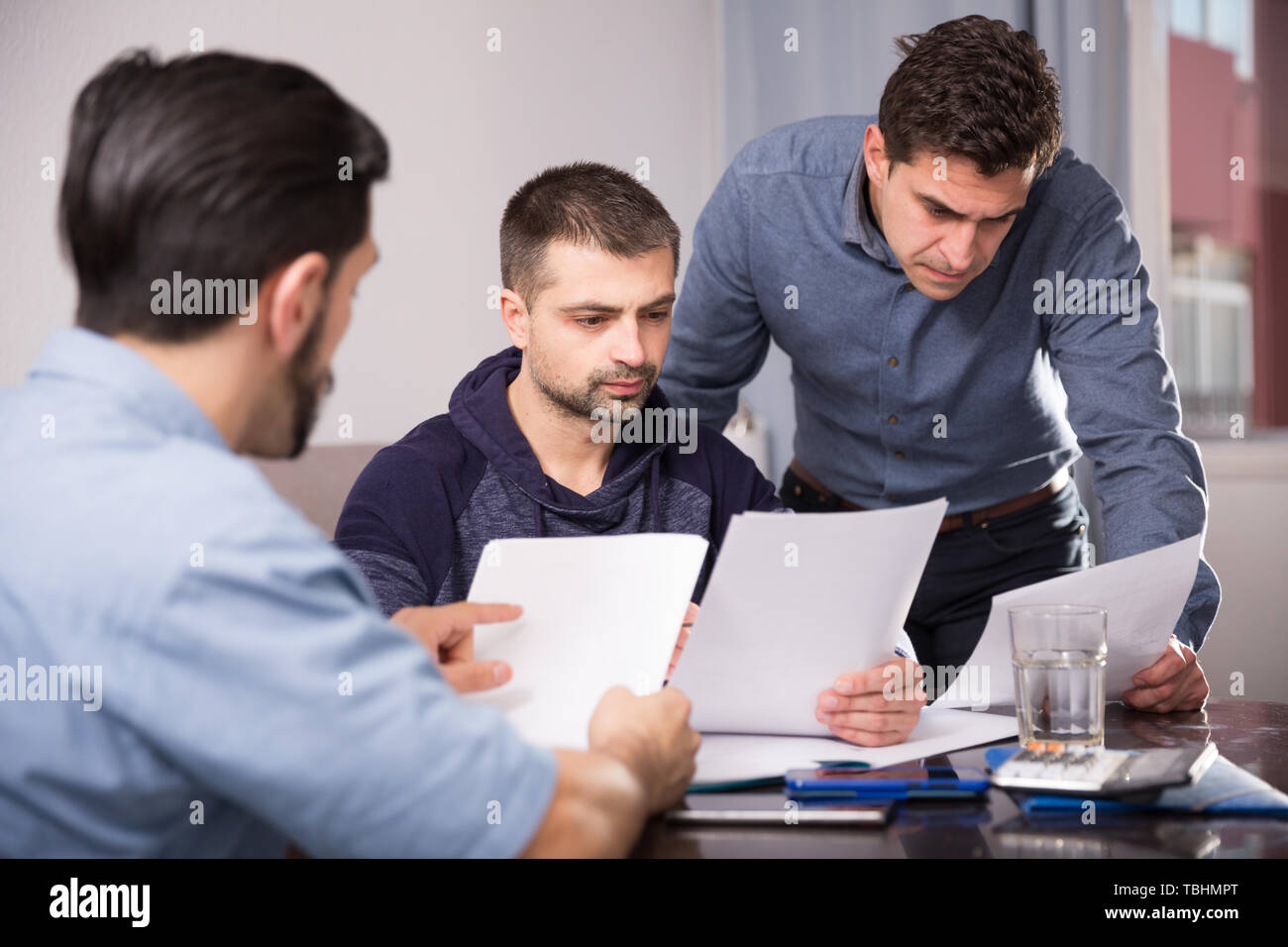 Three upset male friends looking worriedly at papers at home table ...