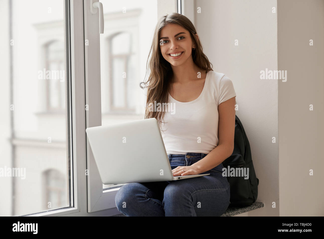Beautiful female student sitting with laptop on windowsill and smiling ...