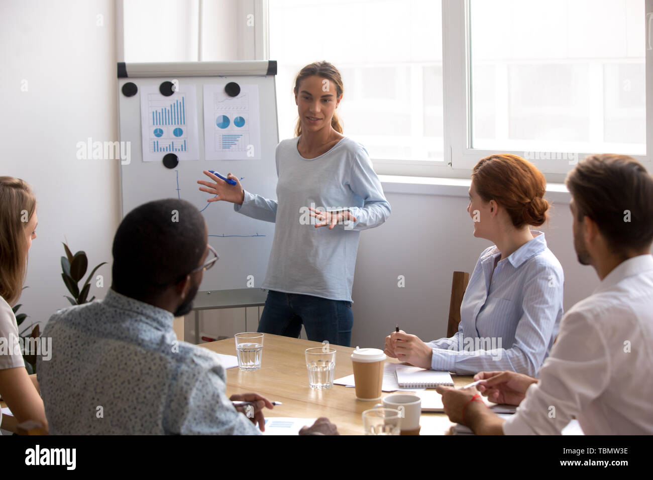 Female business coach standing near whiteboard talking to diverse team ...