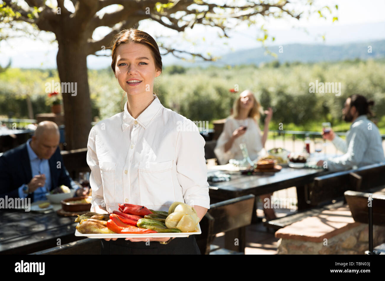 Positive woman waiter demonstrating open-air restaurant to visitors ...
