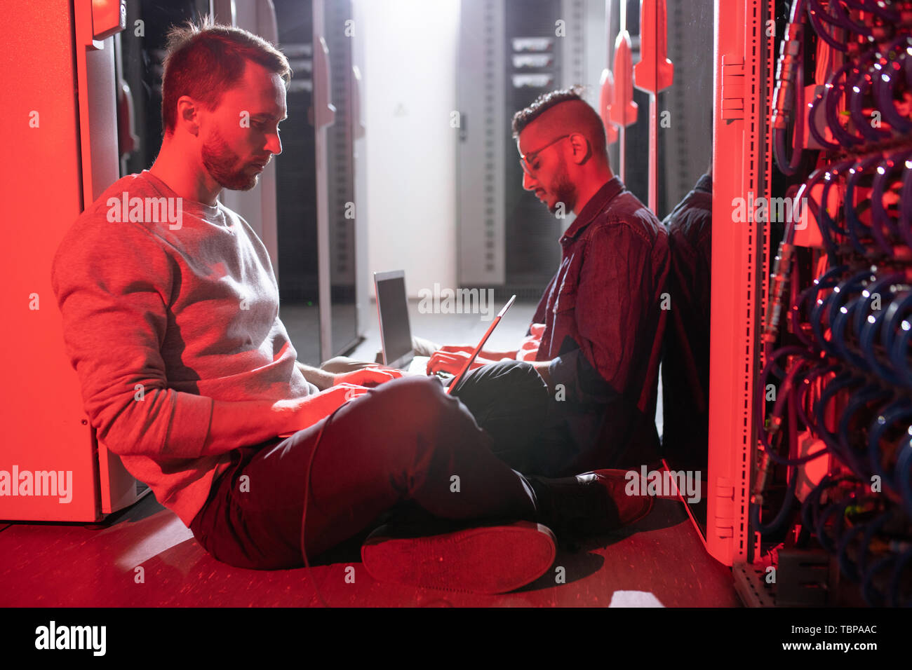 Concentrated young multi-ethnic engineers sitting on floor in server room with red light and ...