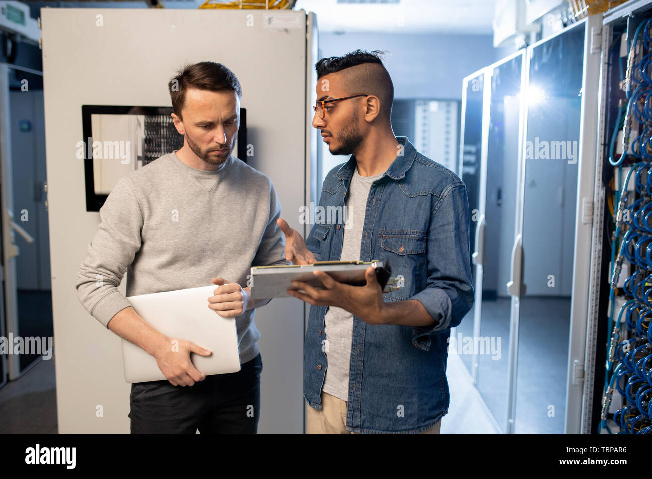 Serious pensive multi-ethnic IT engineers in casual outfits standing in database server room ...