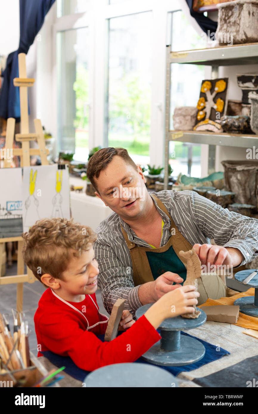 Modeling with teacher. Talented cute boy smiling while modeling clay ...