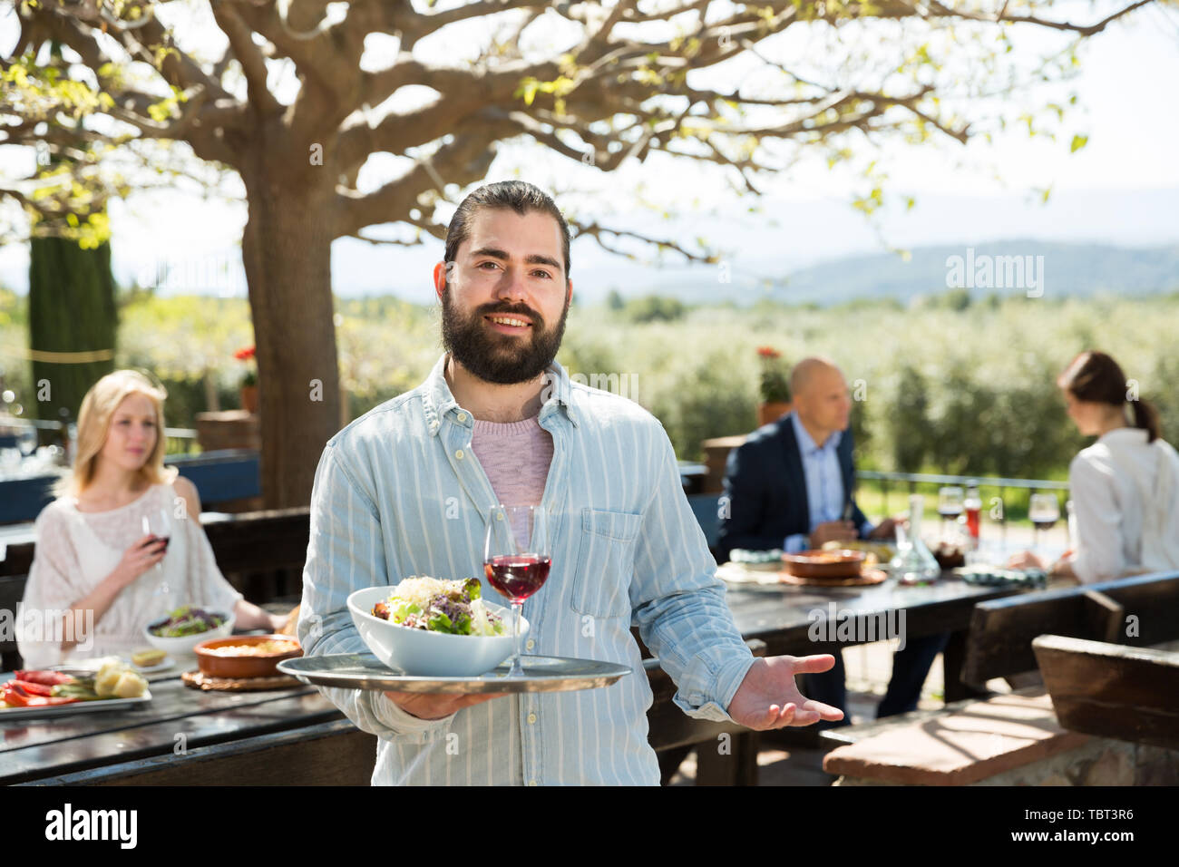 Positive male waiter welcoming guests to country outdoor restaurant ...