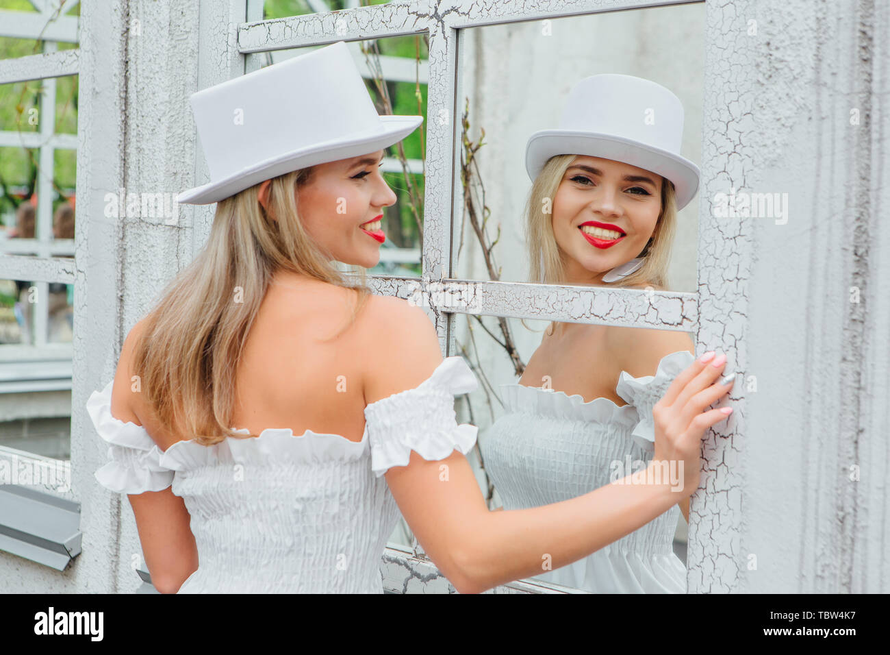 Sexy modern bride in white cylinder hat looks at her reflection in mirror window of an old ...