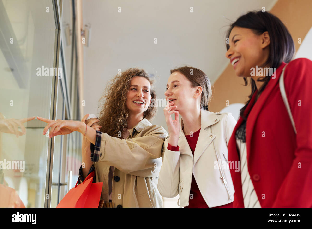 Happy excited young female friends in jackets standing by store window ...