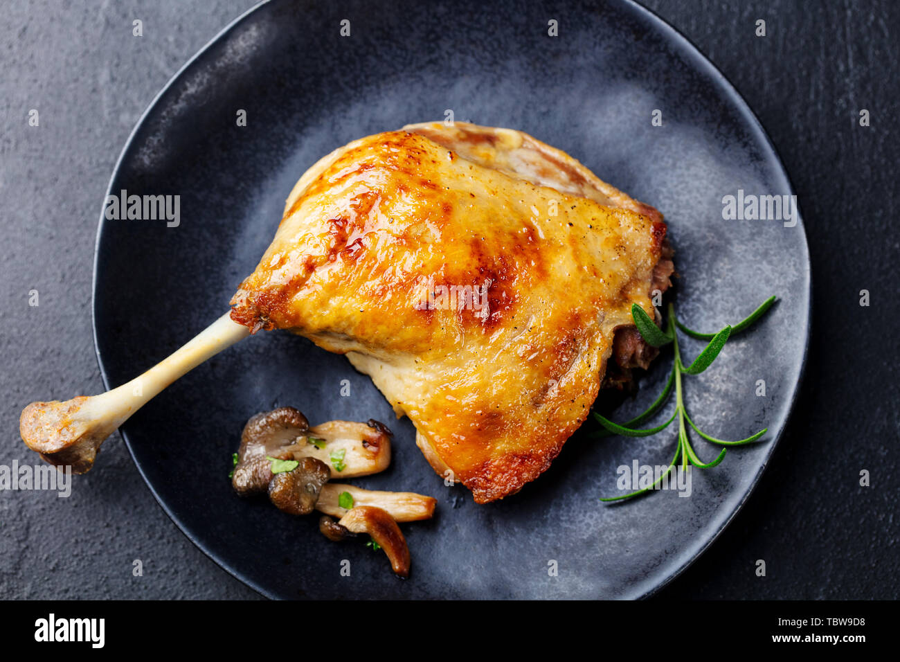 Duck leg confit with mushroom sauce on black plate. Stone background ...