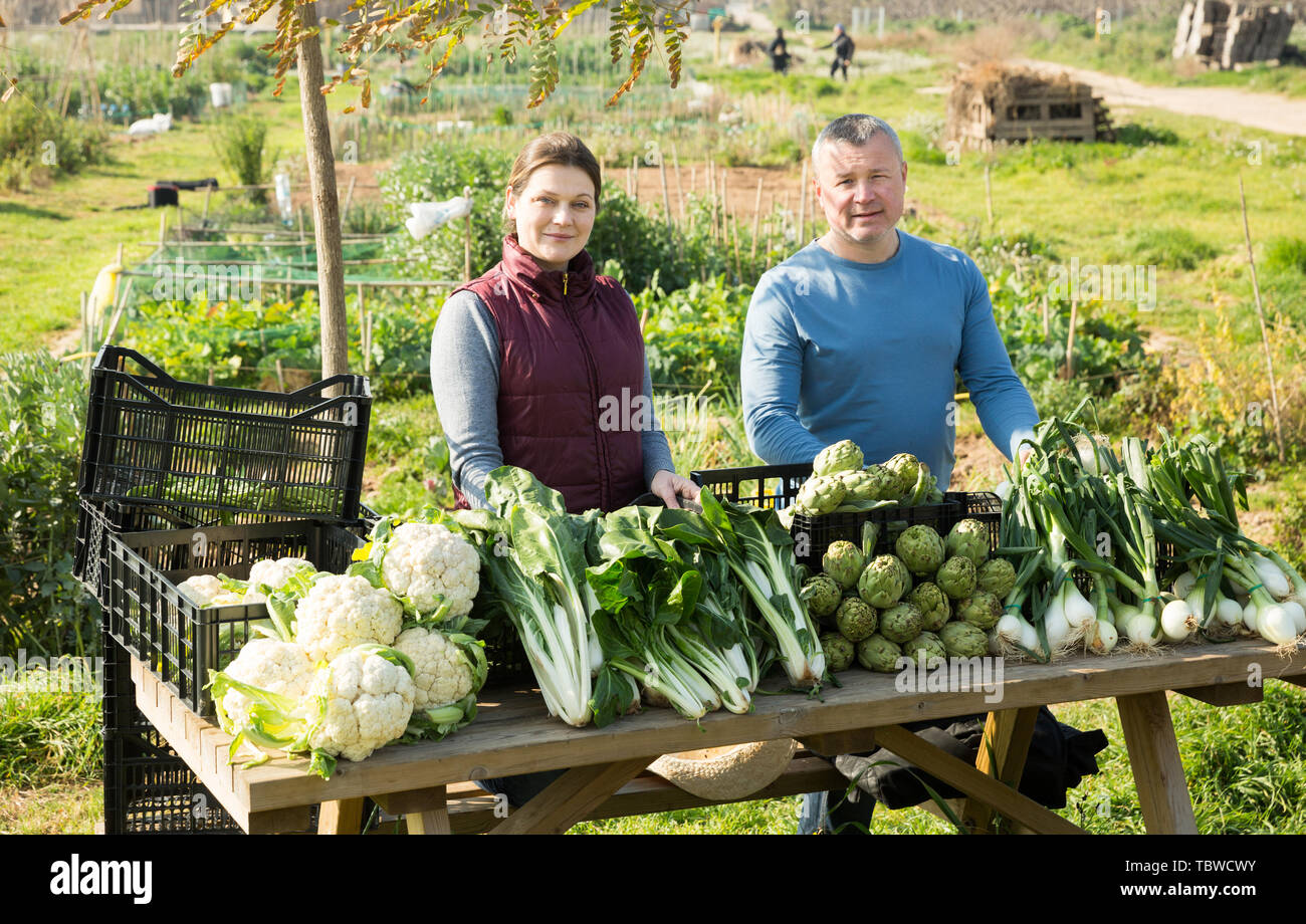 Cheerful couple of farmers standing behind counter with fresh greens ...