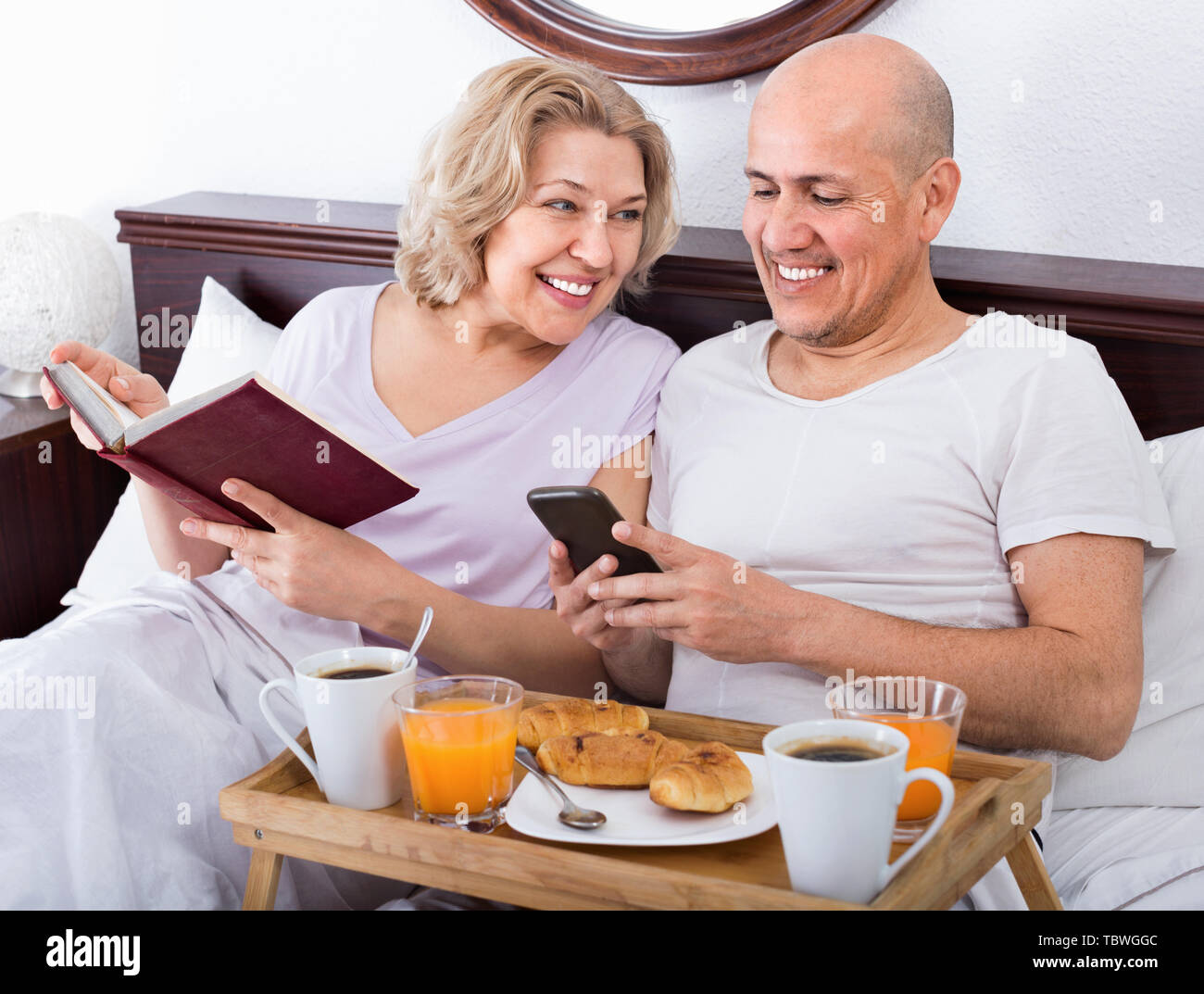 Positive mature adults posing with breakfast and book in bed Stock ...