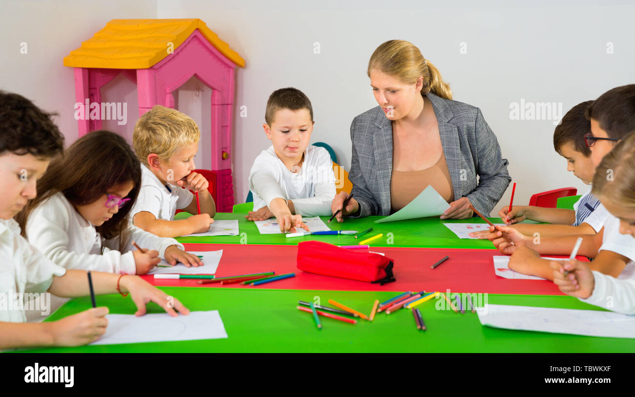 Glad female teacher helping schoolkids drawing with color pencils in ...