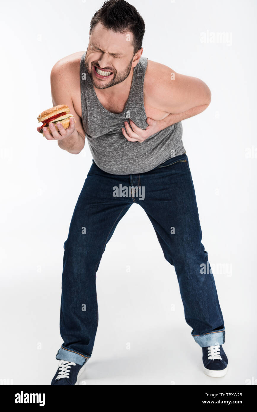 overweight man in tank top holding burger and touching chest on white ...