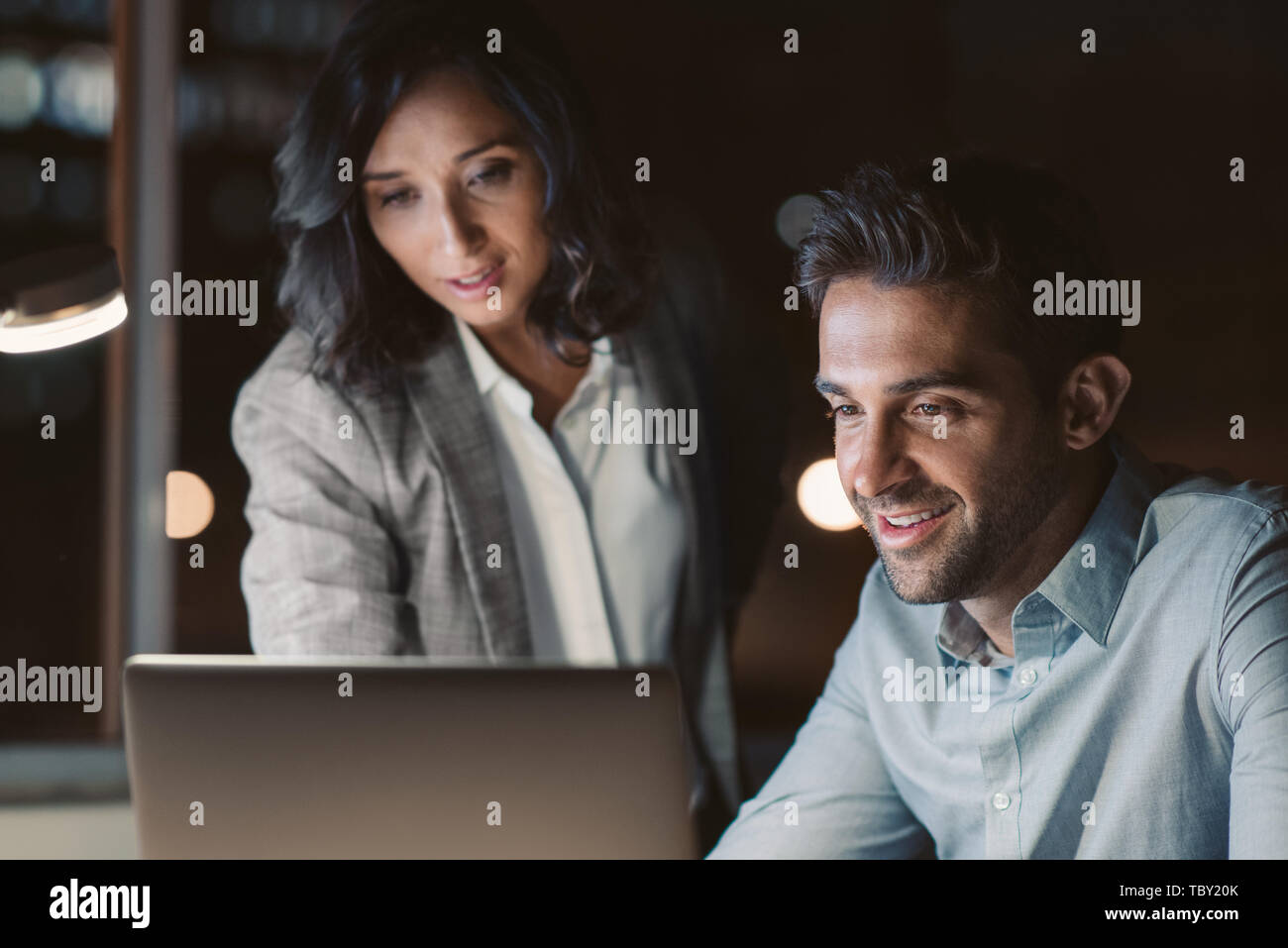Smiling office colleagues using a laptop while working overtime ...
