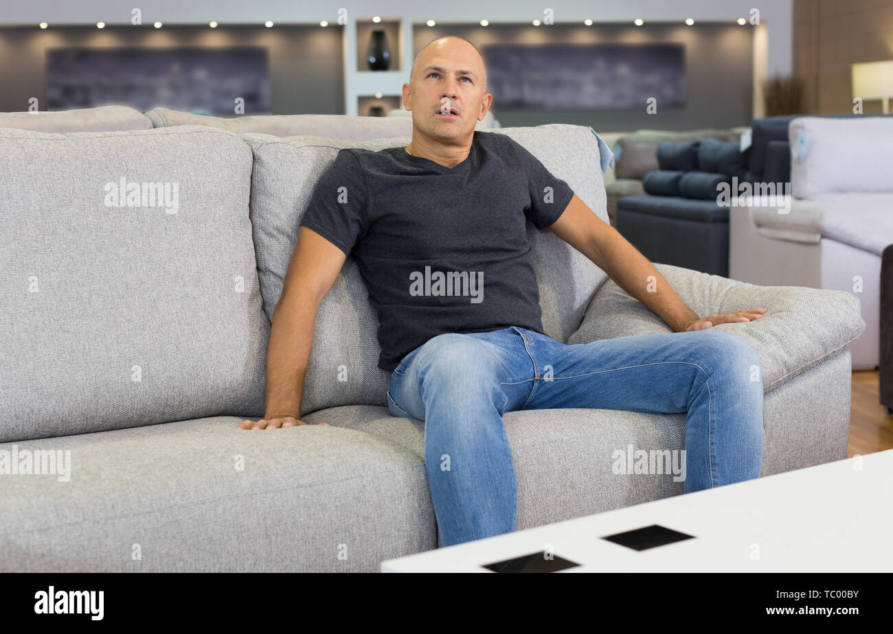 Portrait of positive man testing light gray sofa in furniture store ...