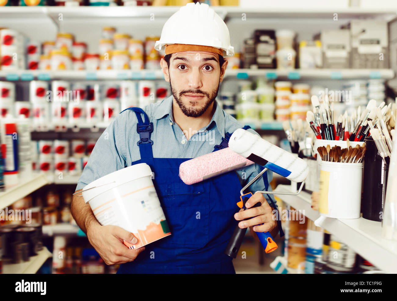 Portrait of young workman holding bucket of paint and rollers in paint ...