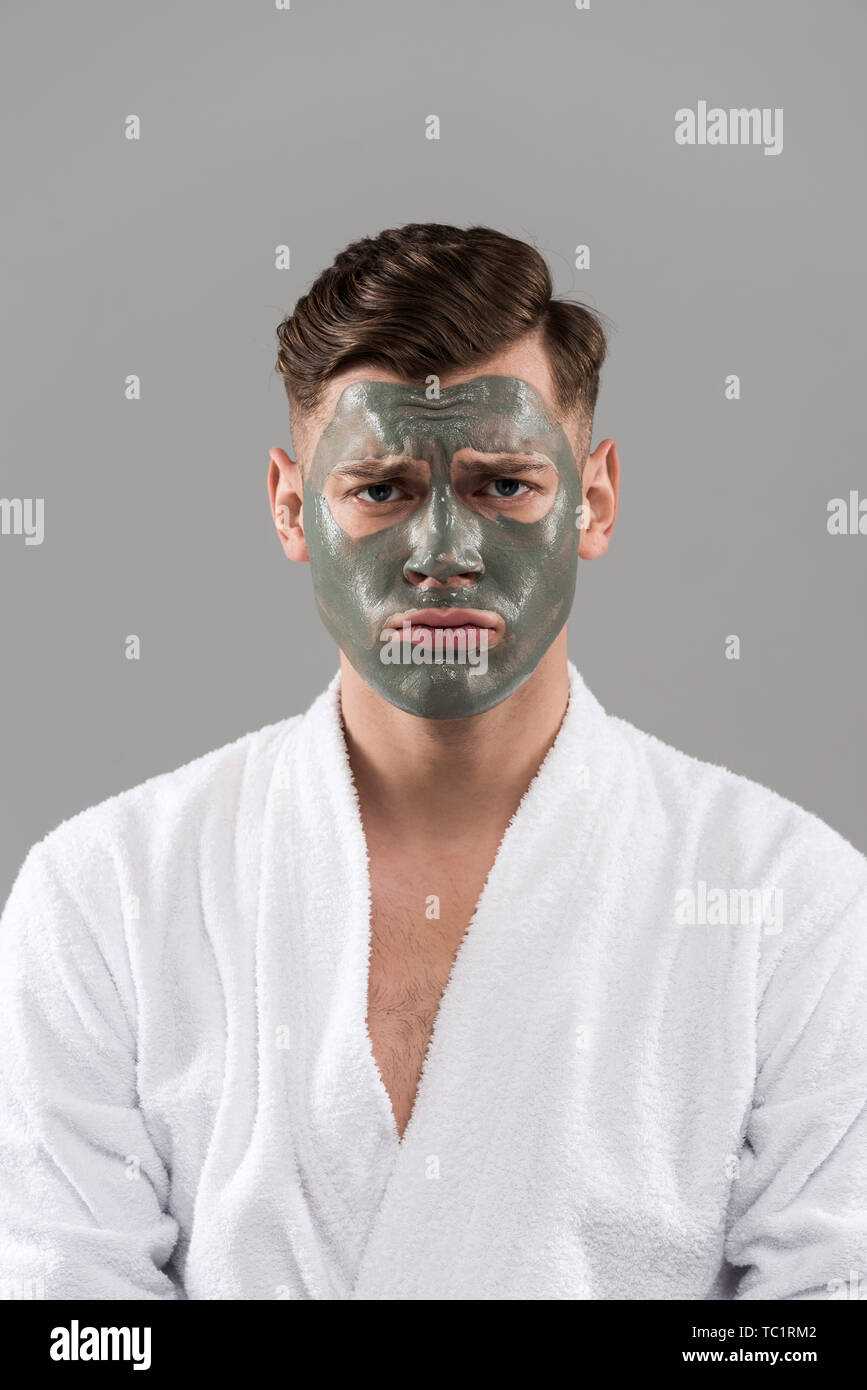front view of sad young man in bathrobe with clay mask isolated on grey ...