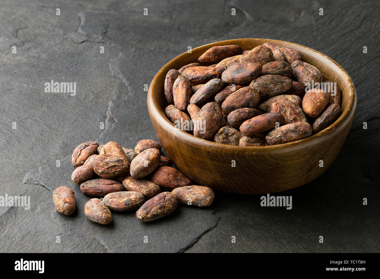 Roasted unpeeled cocoa beans in a brown wooden bowl next to a pile of ...