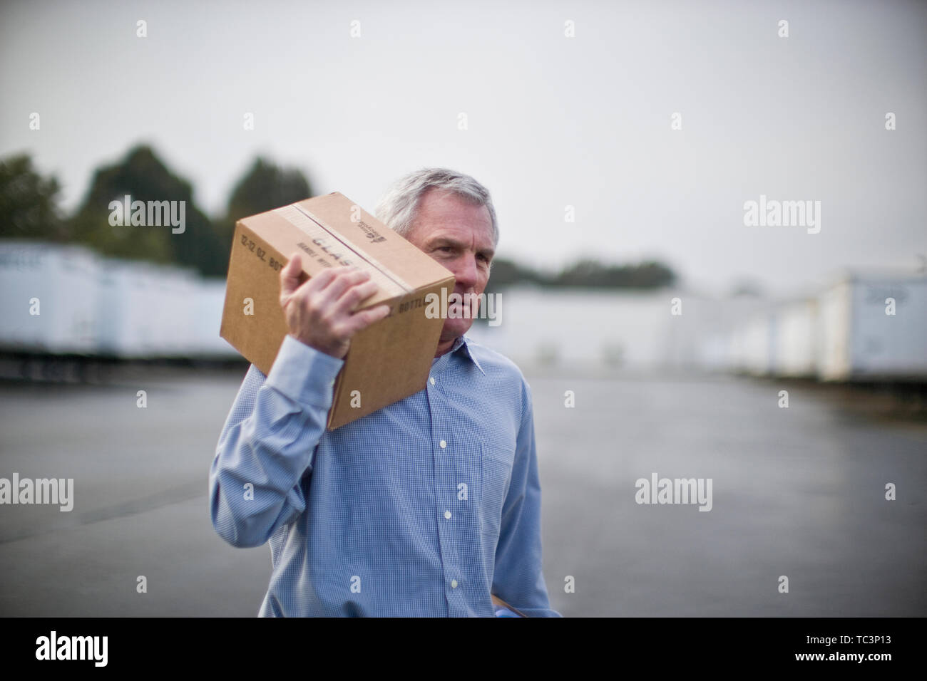 Mature man carrying a cardboard box on his shoulder at a shipping yard ...