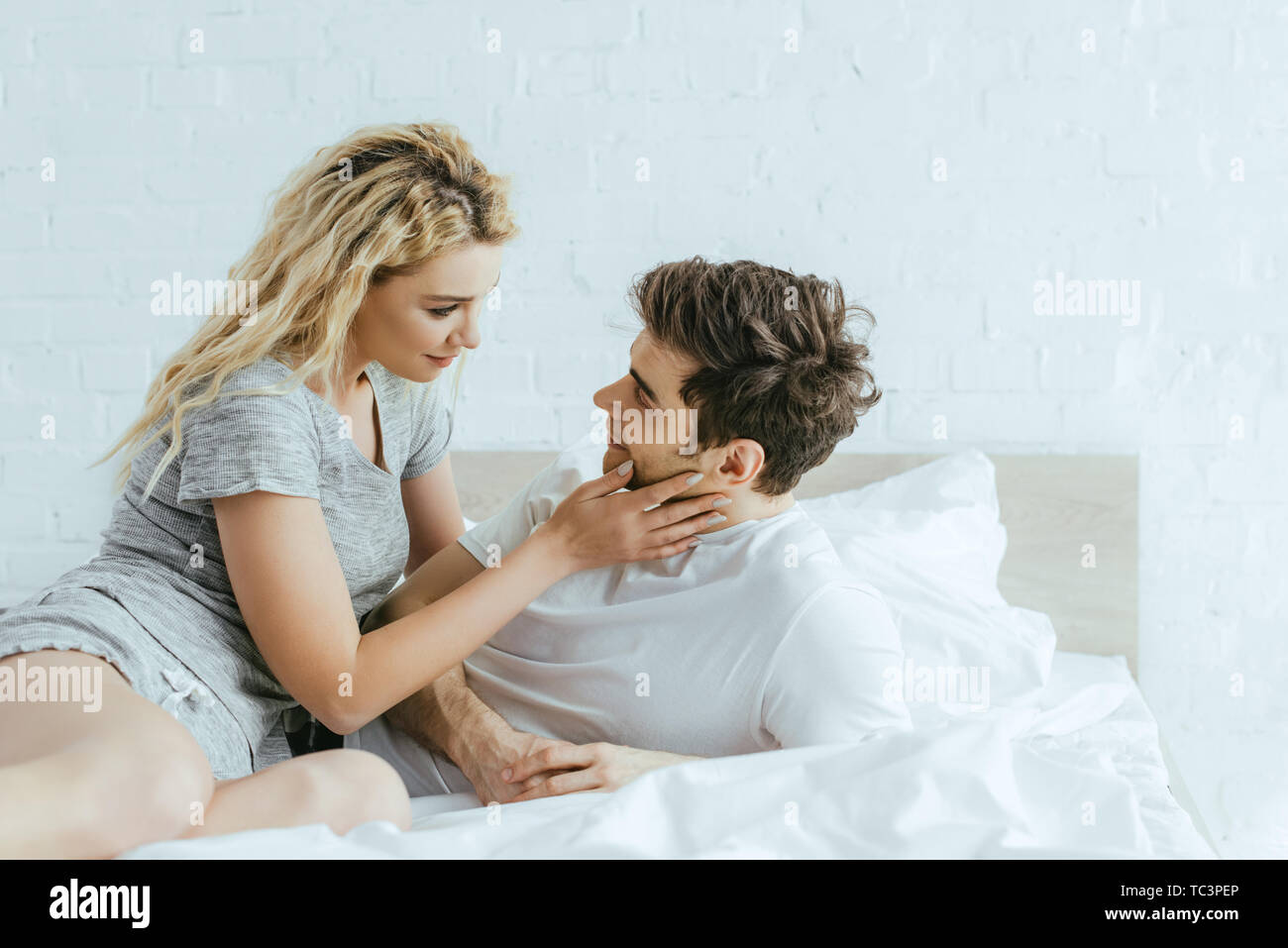 attractive blonde woman touching cheek of handsome man lying on bed ...