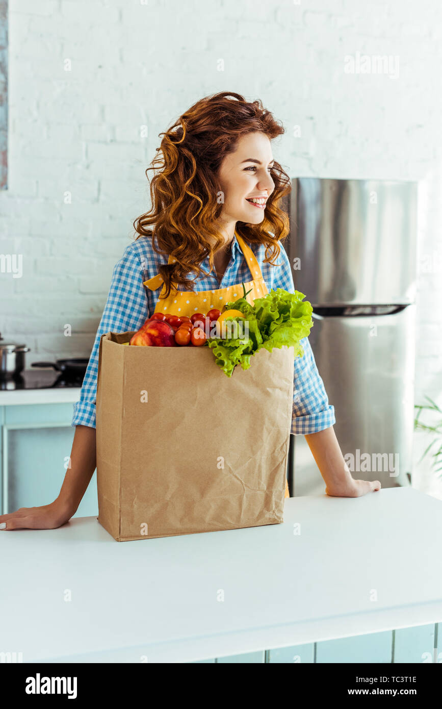 smiling woman standing near paper bag with ripe fruits and vegetables in kitchen Stock Photo - Alamy