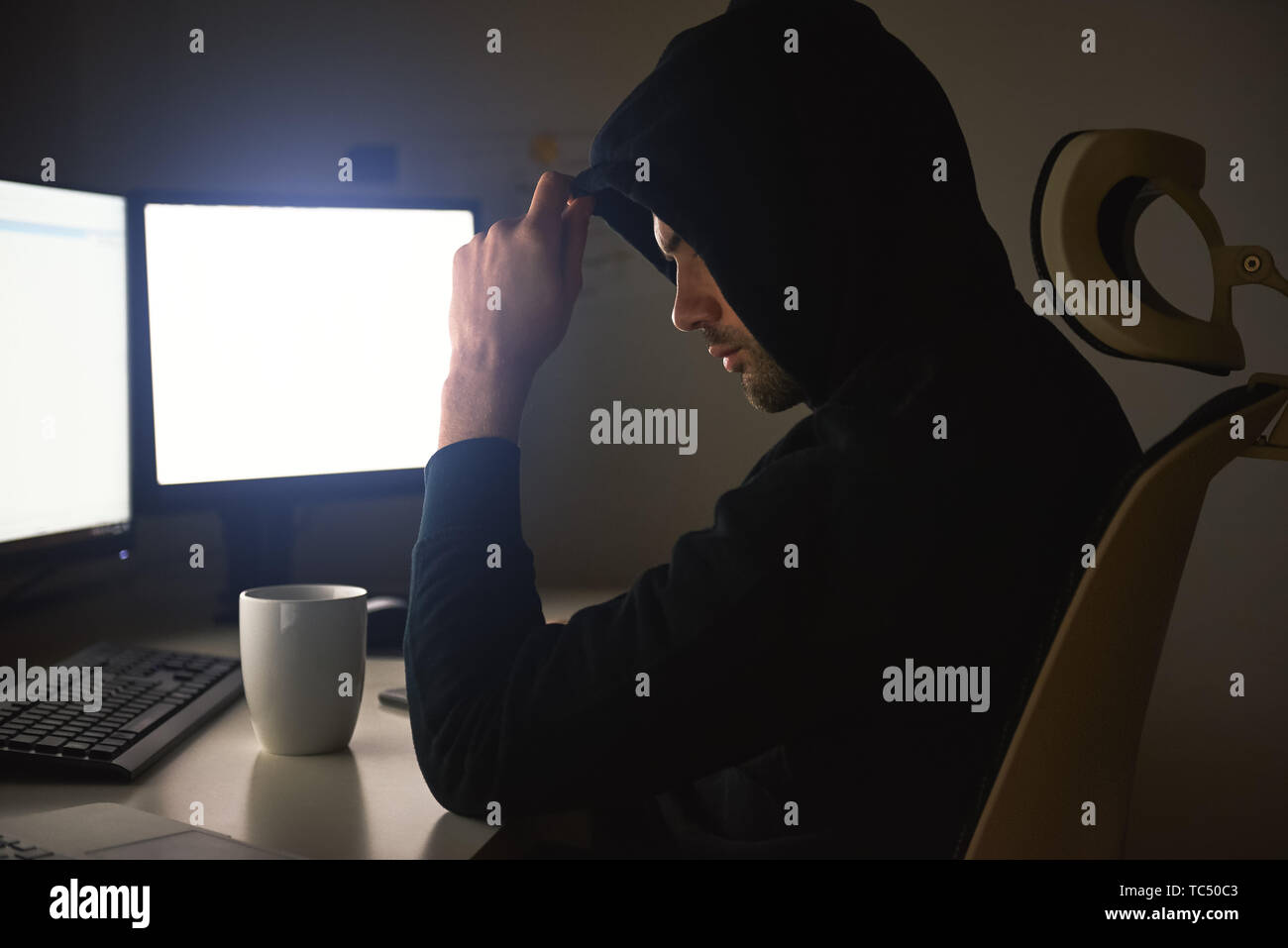 Computer hacker. Young man in black hoodie using multiple computers for stealing data while ...