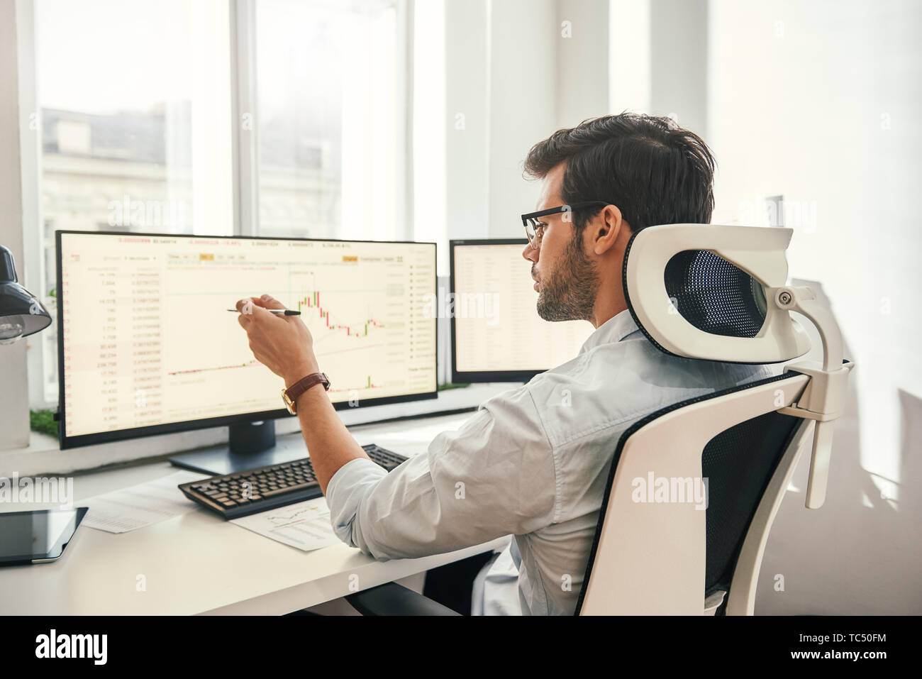 Analyzing data. Back view of young businessman or trader pointing on the data on computer screen ...