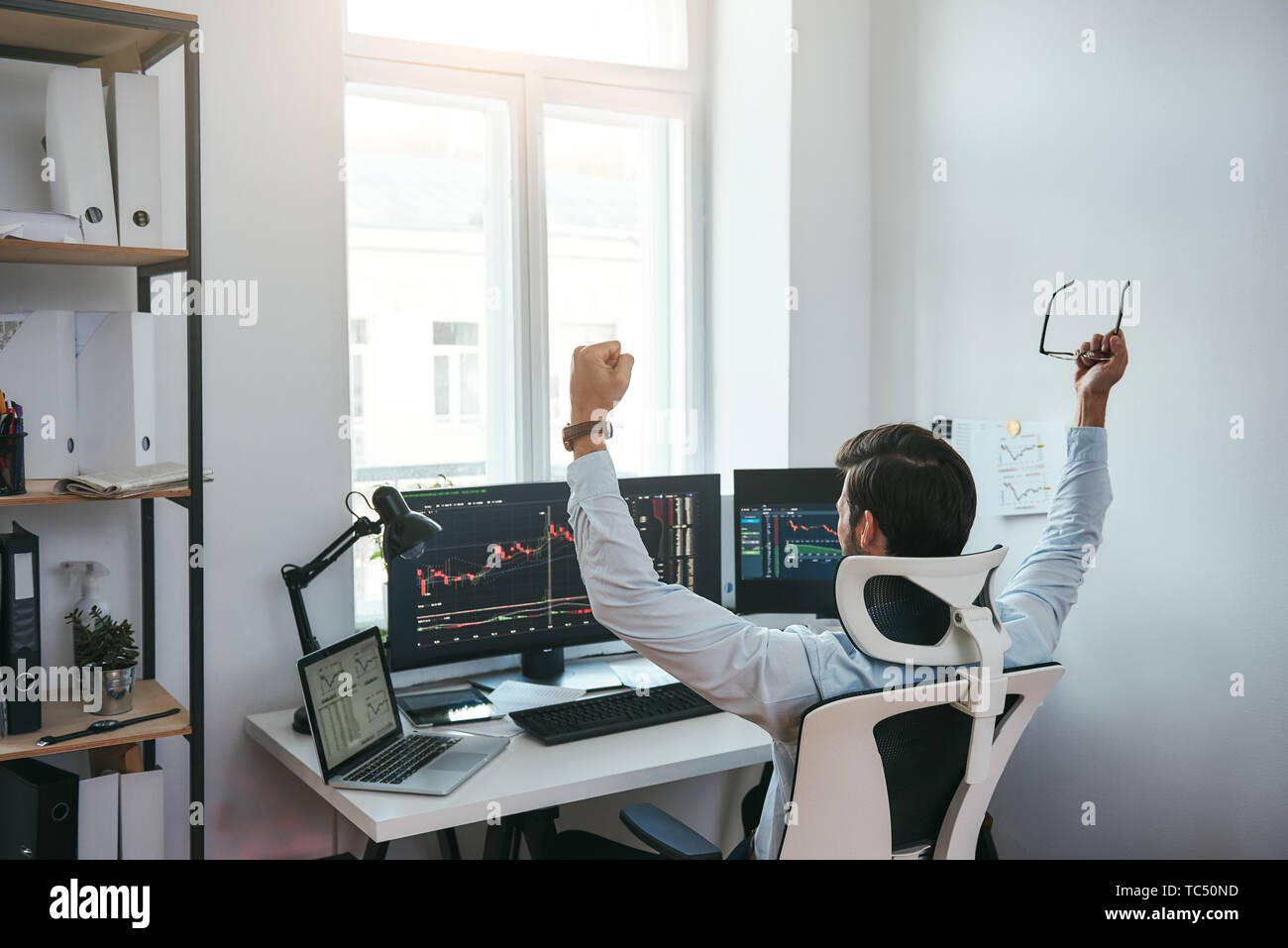 Winner Back view of stock trader with raised hands holding eyeglasses and feeling happy while ...