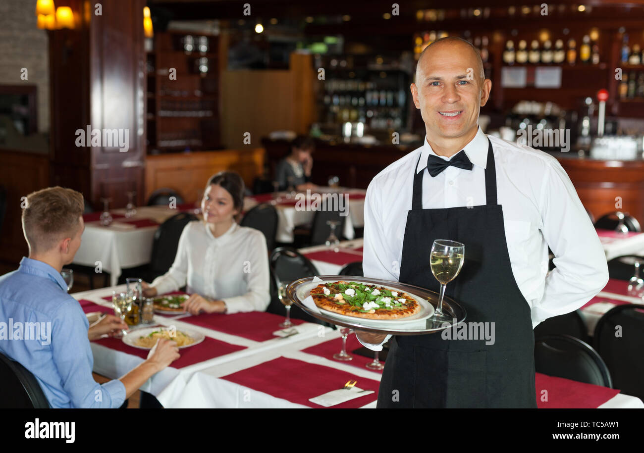 Polite waiter holding tray in restaurant with customers behind him ...