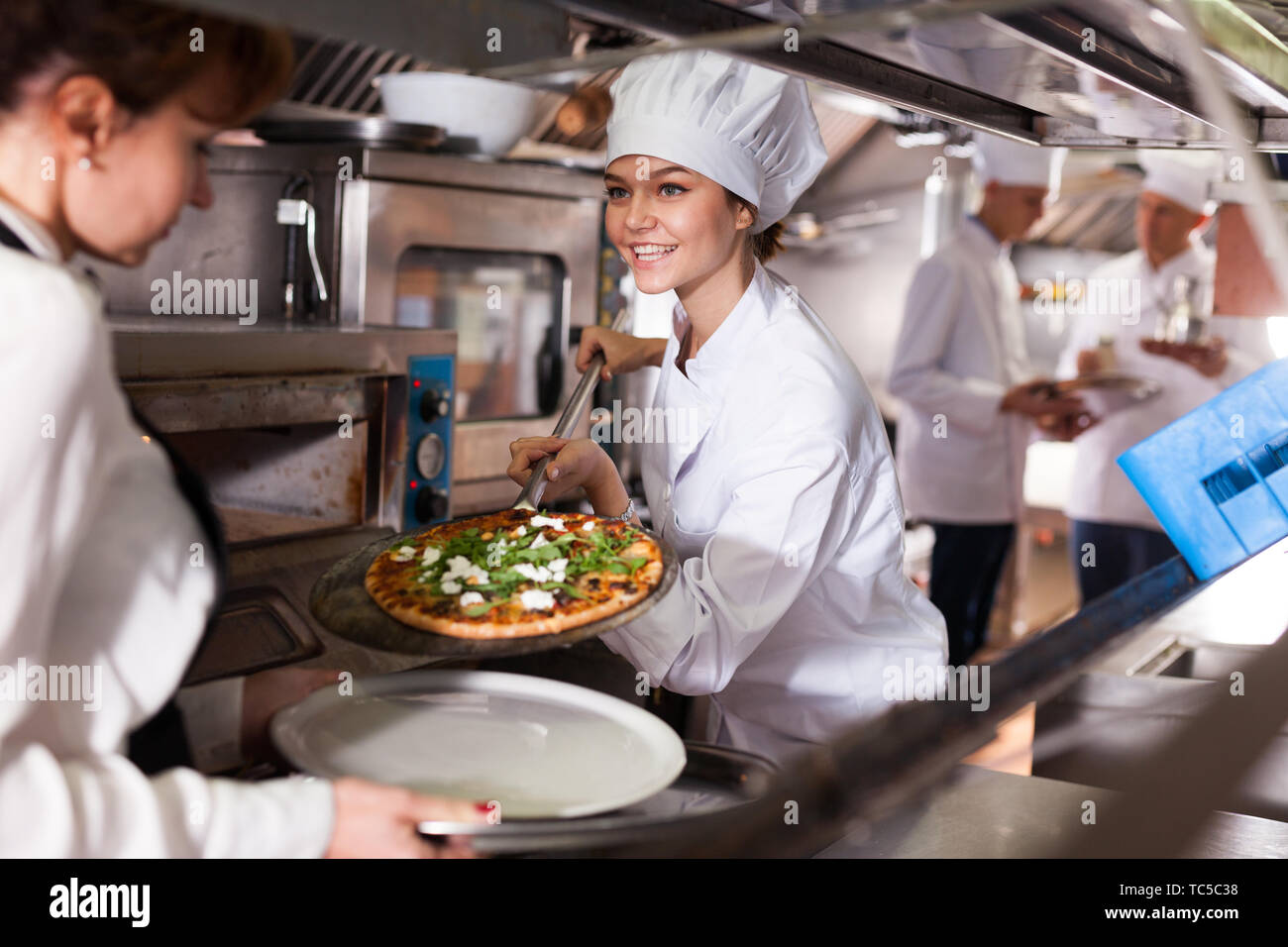 Smiling young female chef giving cooked pizza to waitress ready to ...