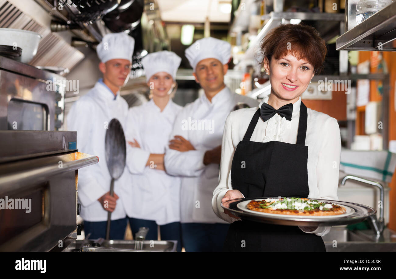 Smiling waitress standing in restaurant kitchen with ordered pizza ...