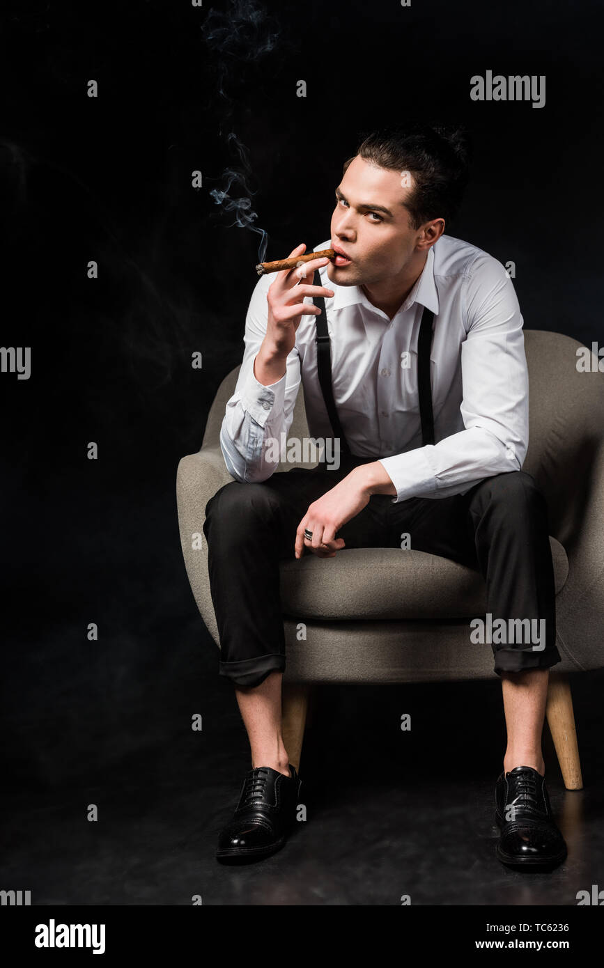 serious man smoking cigar while sitting in armchair on black Stock ...