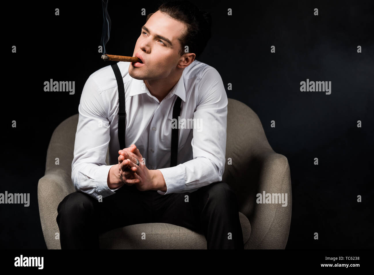 serious man smoking cigar while sitting in armchair with clenched hands ...