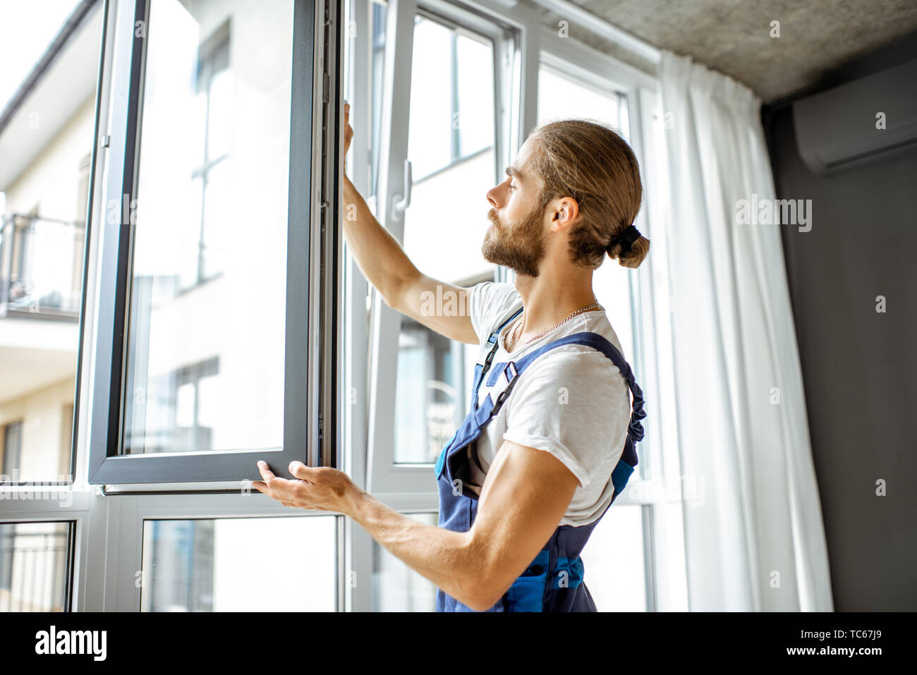 Workman in overalls installing or adjusting plastic windows in the living room at home Stock ...