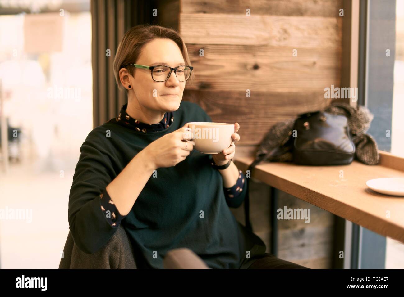 smirking woman with coffee cup sitting indoors in café, in Munich ...