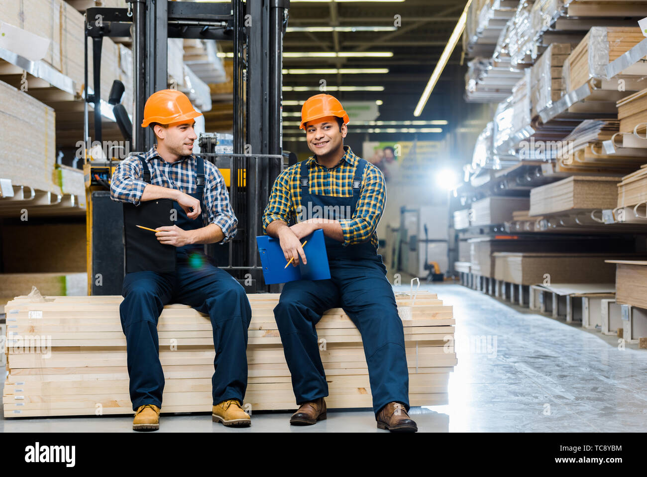 two smiling multicultural workers with clipboards sitting on plywood ...