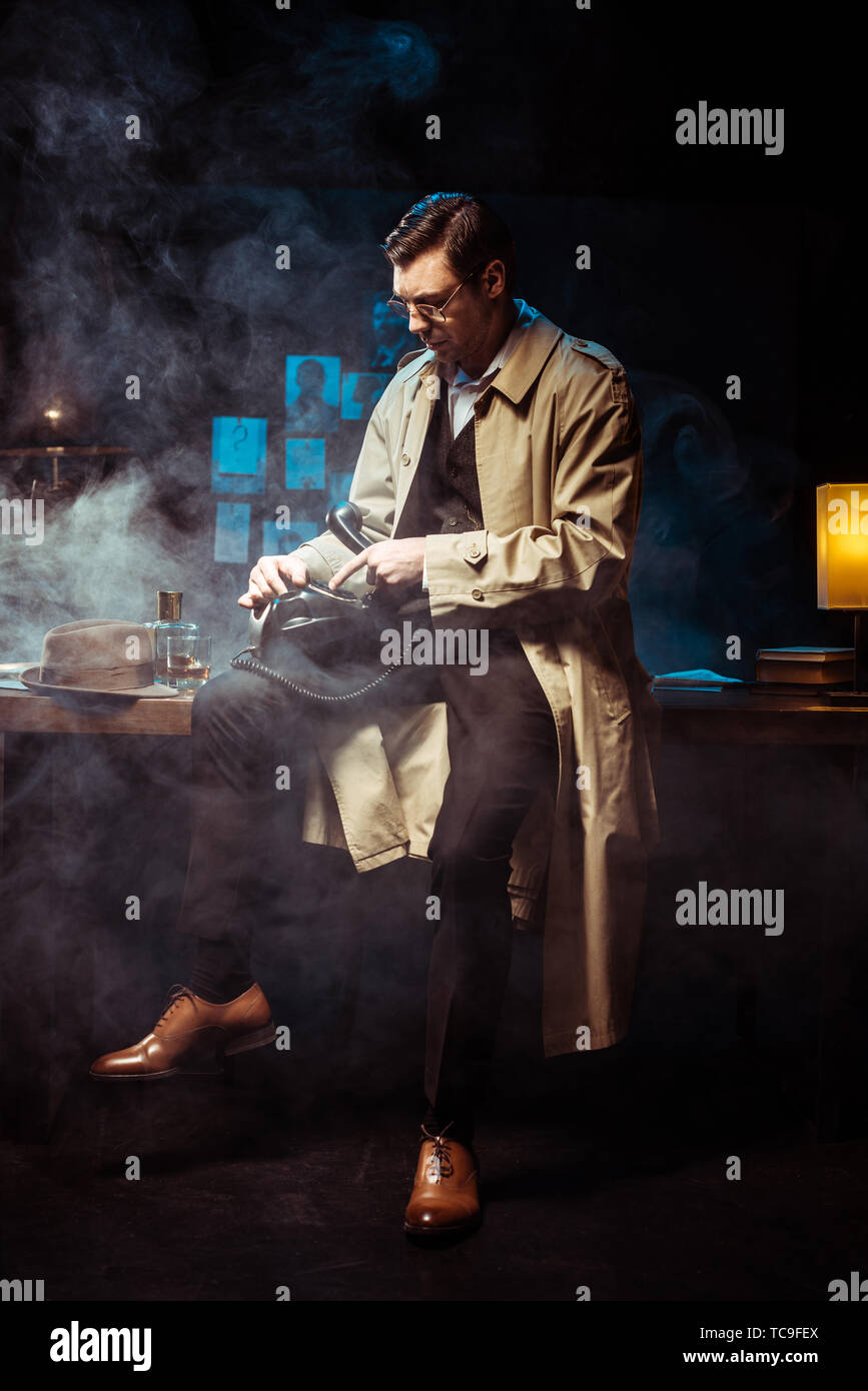 Detective in trench coat sitting on table and using telephone in dark ...