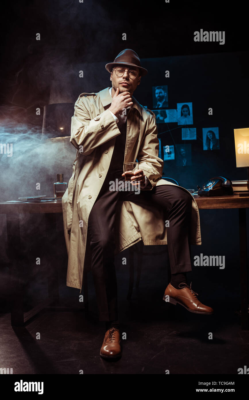 Detective in trench coat and hat sitting on table and holding glass of ...