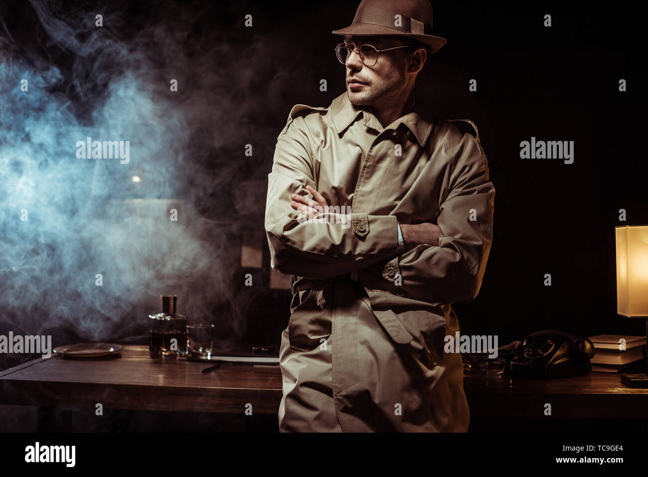 Detective in trench coat and hat standing with crossed arms in office ...