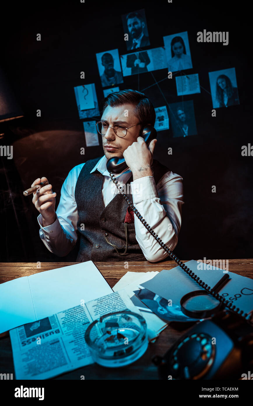 Detective in glasses holding cigar and talking on telephone in dark ...