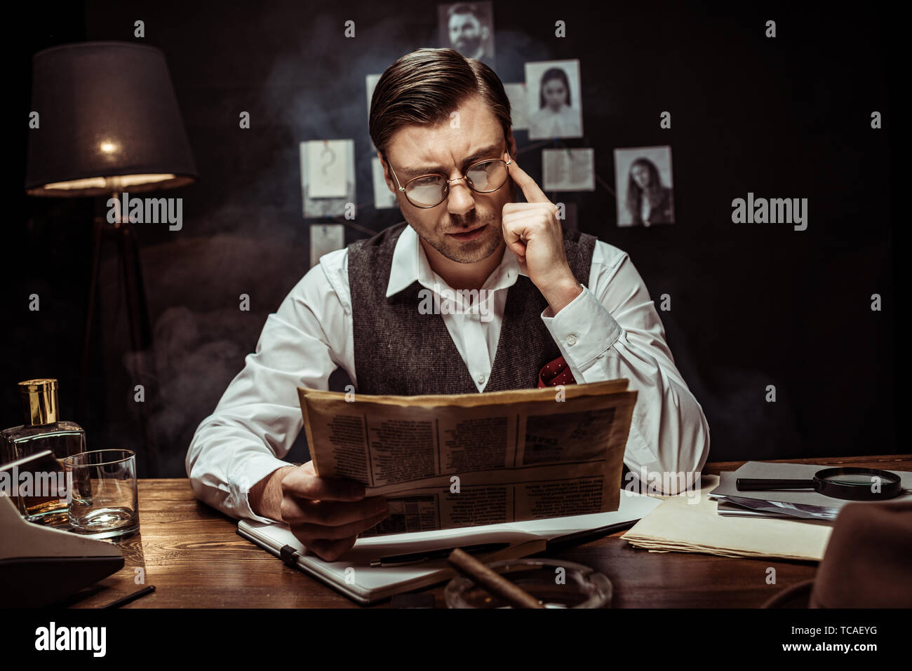 Concentrated detective in glasses reading newspaper in dark office ...