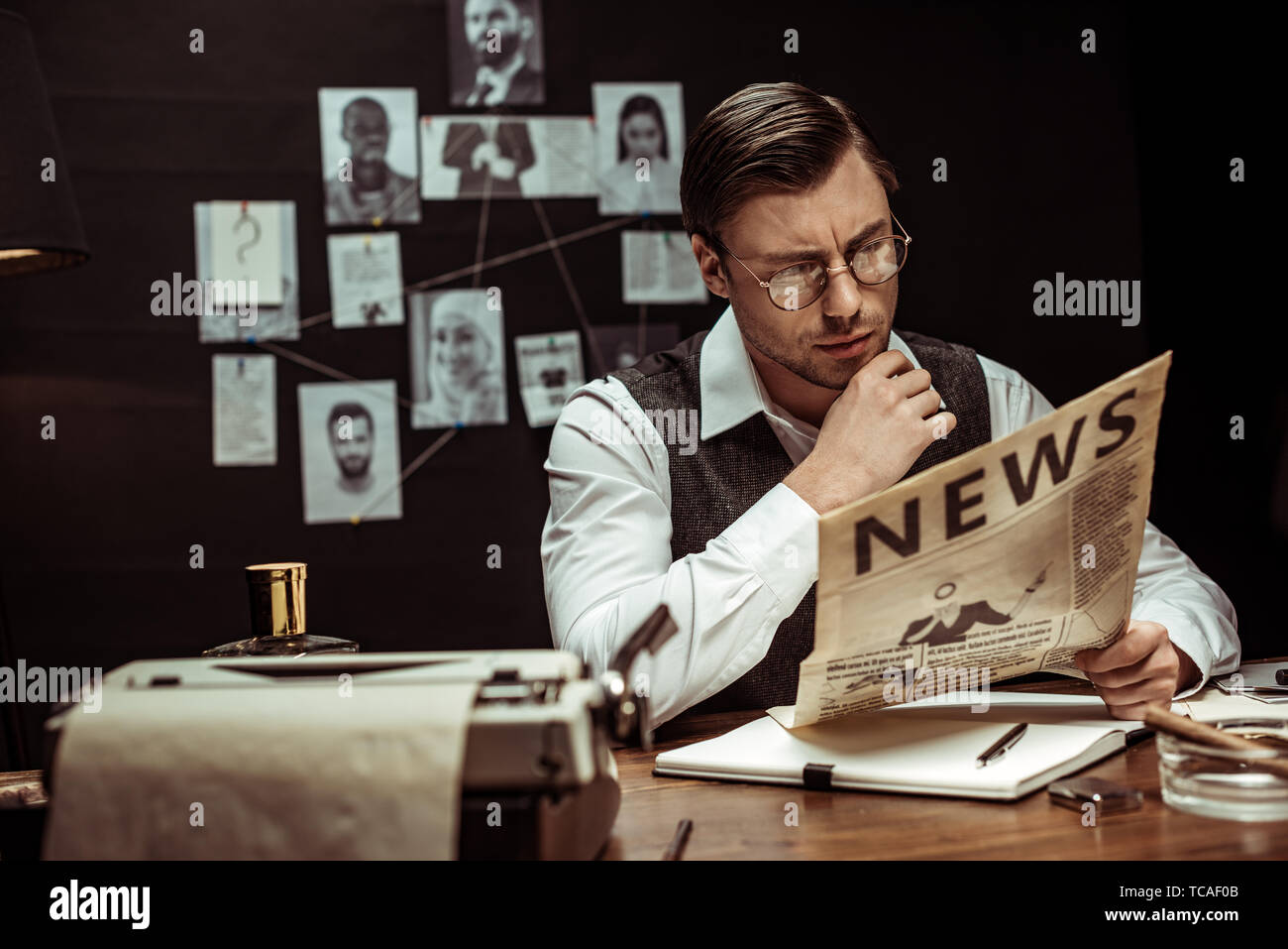 Concentrated detective in glasses reading newspaper in dark office ...