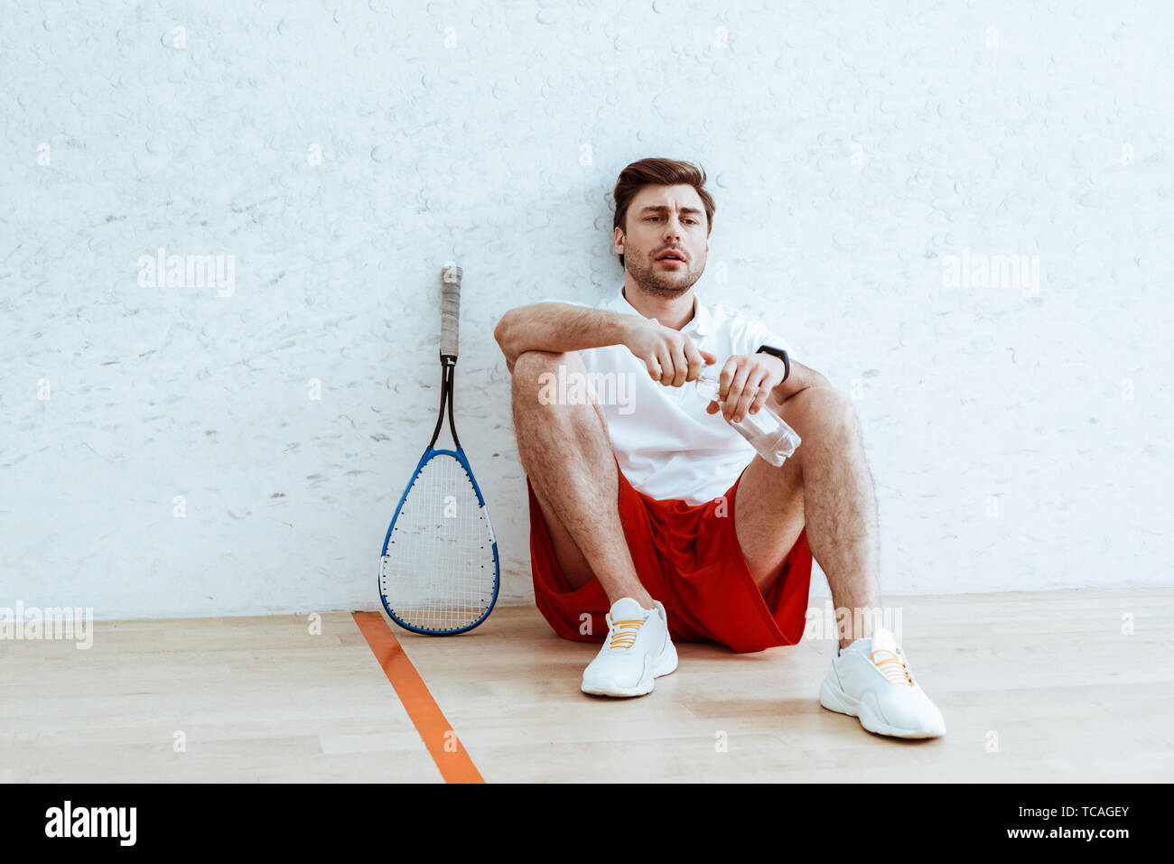 Tired squash player sitting on floor and opening bottle of water Stock ...