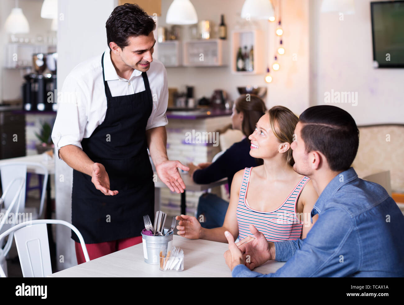 Portrait of smiling cafe waiter standing at table and talking to guests ...