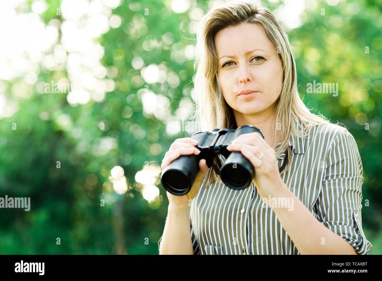A blond woman with black binoculars stays outdoor, green out of focus ...