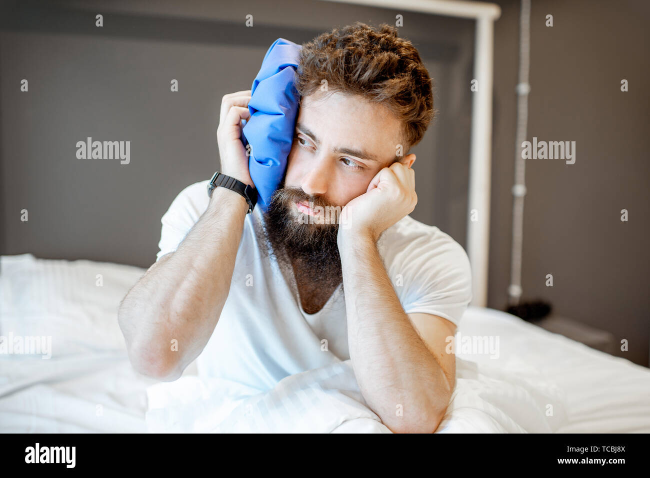Man feeling bad, sitting on the bed with headache and applying ice bag ...