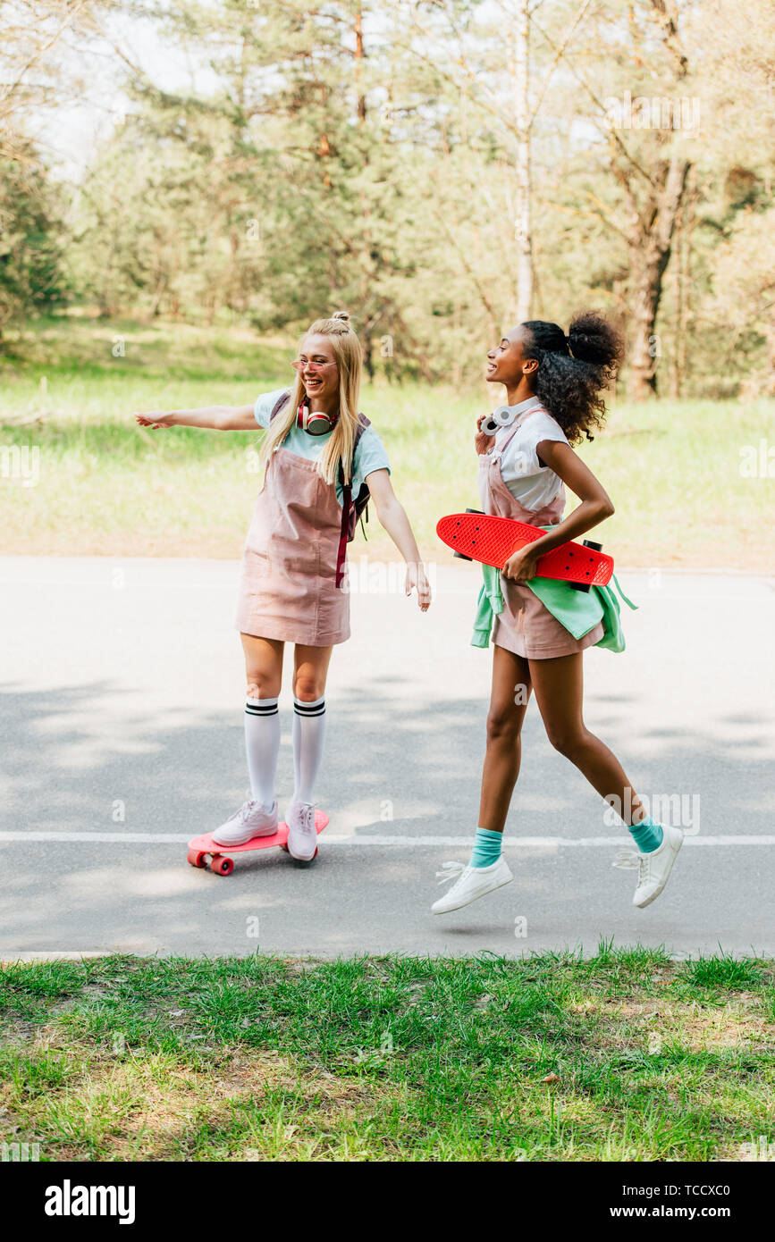full length view of two multicultural friends skateboarding on road ...